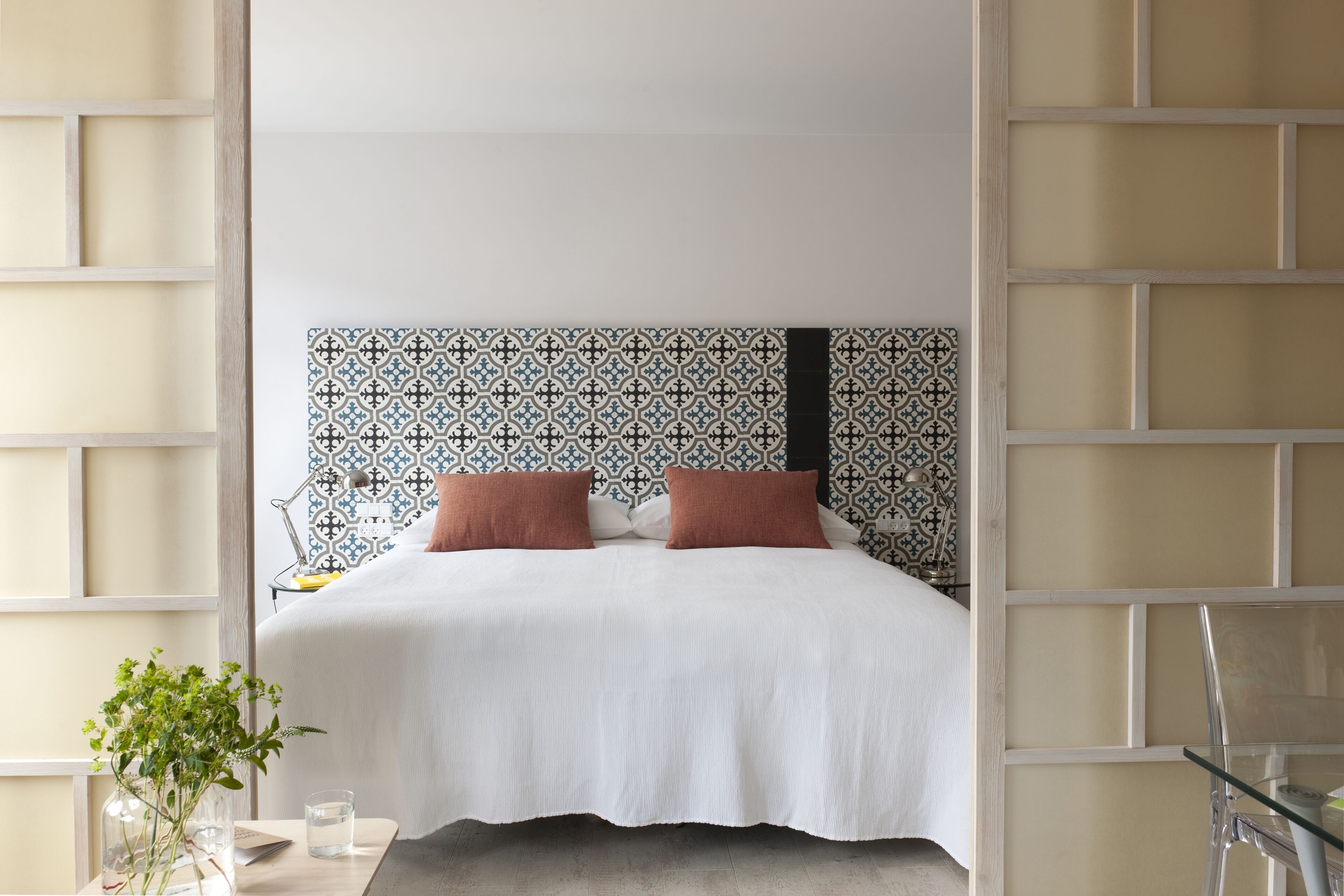 Quarto moderno com cabeceira estampada, cama com roupa de cama branca e duas almofadas, mesa de cabeceira com abajur e mesa de vidro com vaso e copo com água.