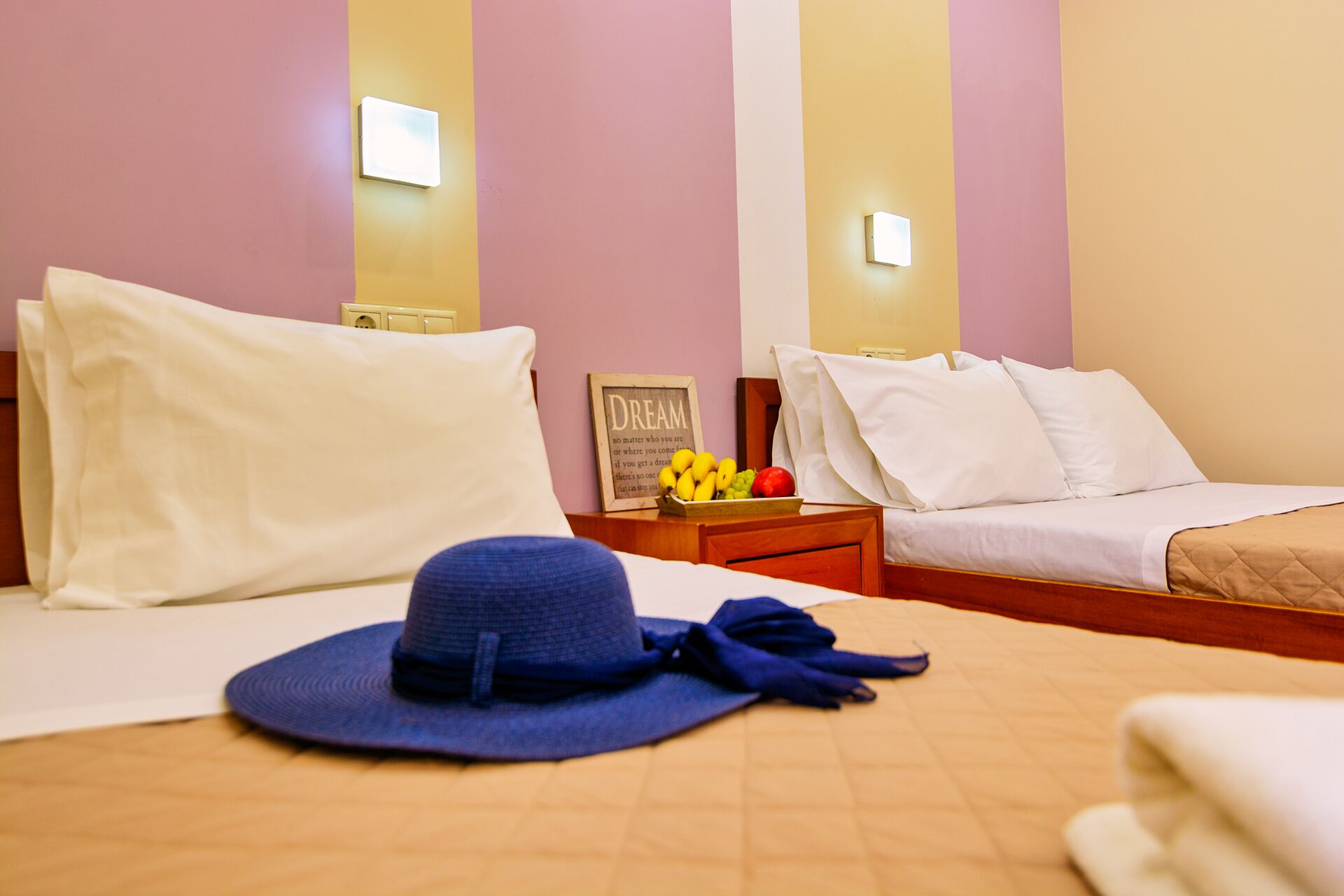 Quarto de hotel com duas camas, uma mesa de cabeceira com uma fruteira e um candeeiro fixado na parede.