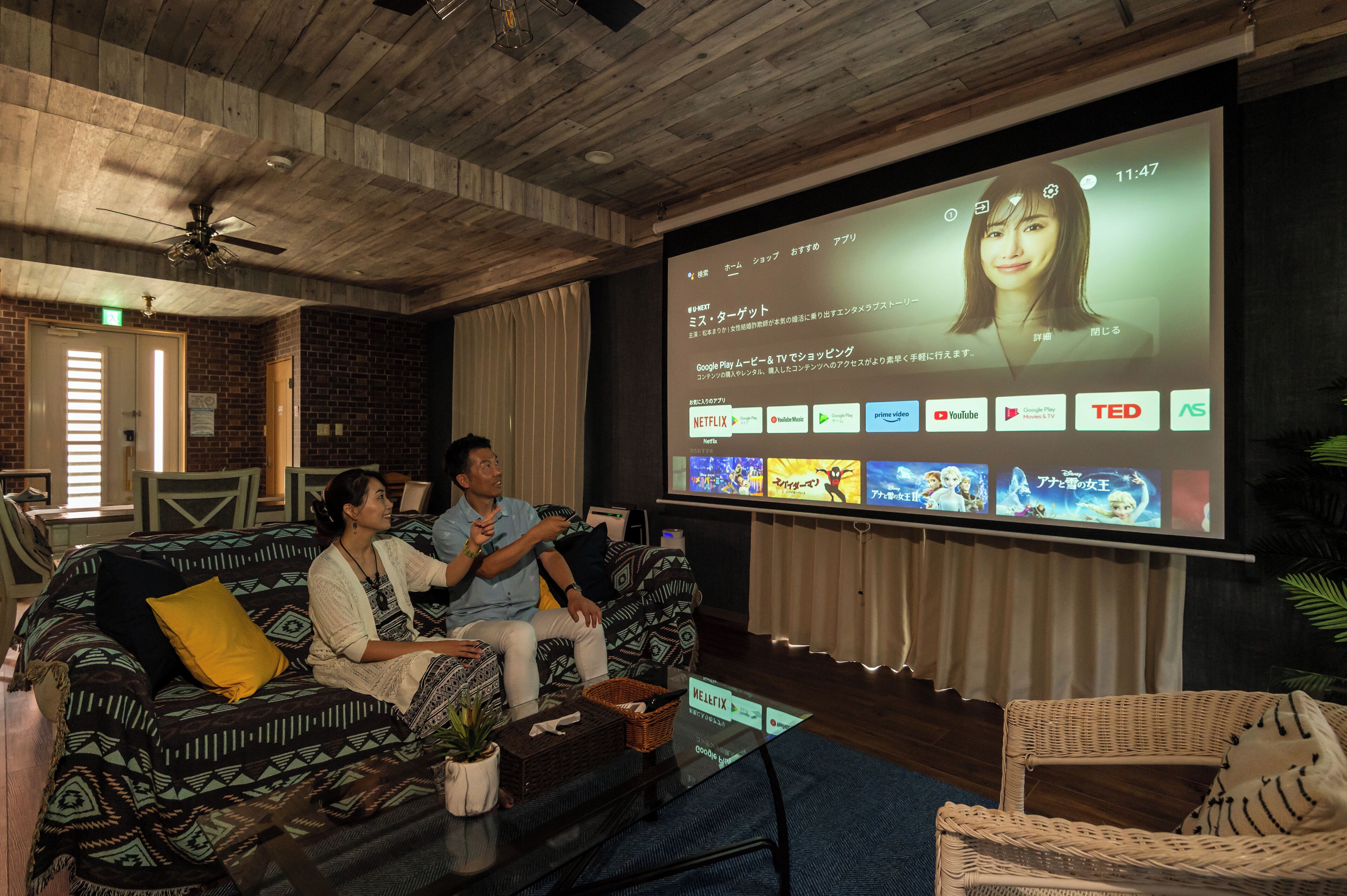 Uma sala de estar moderna com uma tela grande exibindo diversos serviços de streaming, um ventilador de teto e um espaço de estar aconchegante com almofadas estampadas.