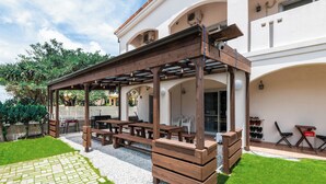 Uma área de estar coberta ao ar livre, com bancos e mesas de madeira, estrutura de pérgola e um caminho de cascalho que leva a um edifício com fachada branca e janelas em arco.