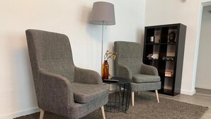 Uma sala de estar com dois poltronas cinzas, uma mesa redonda pequena, um abajur e uma estante com objetos decorativos.
