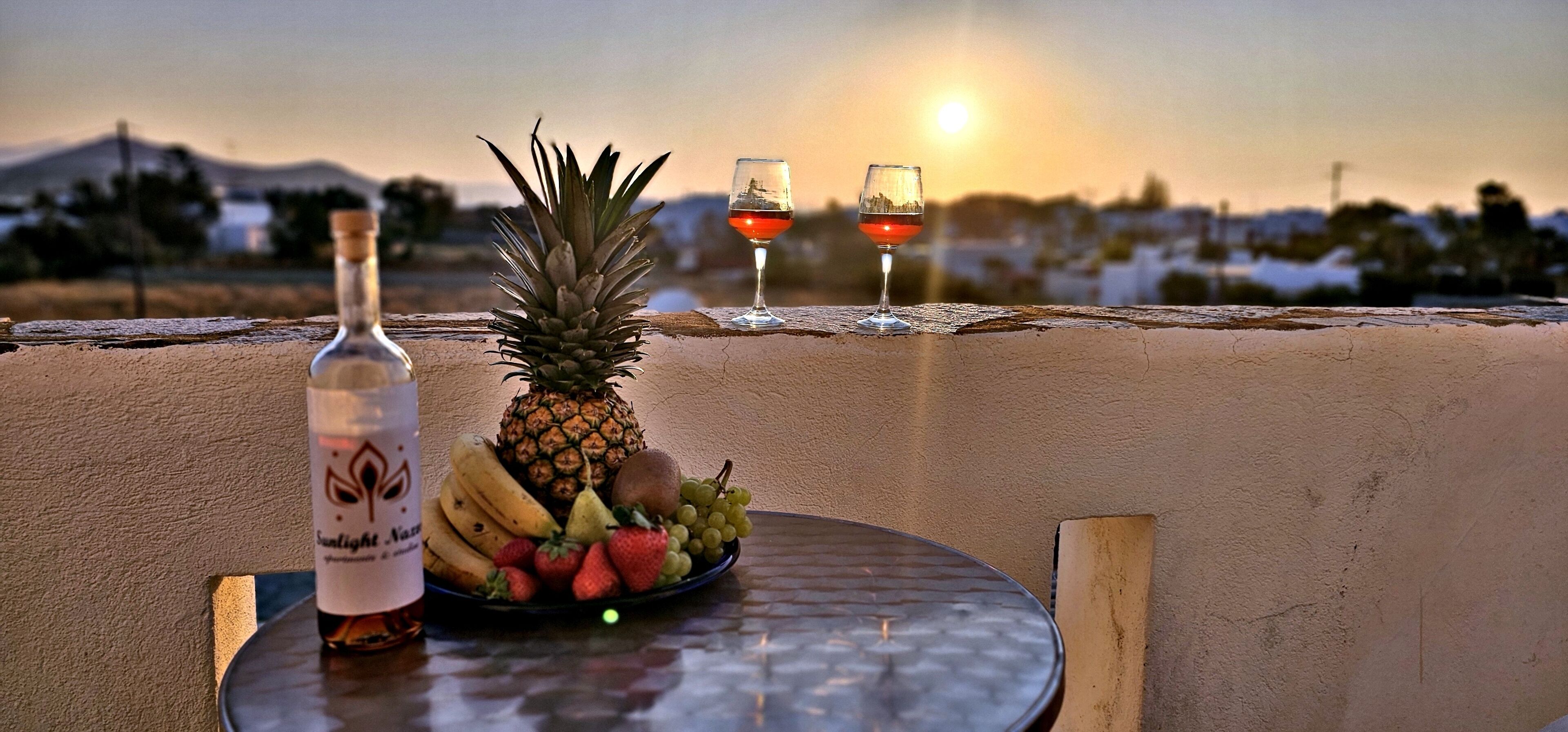 Uma mesa com uma garrafa de vinho, uma abacaxi e um prato de frutas ao pôr do sol.