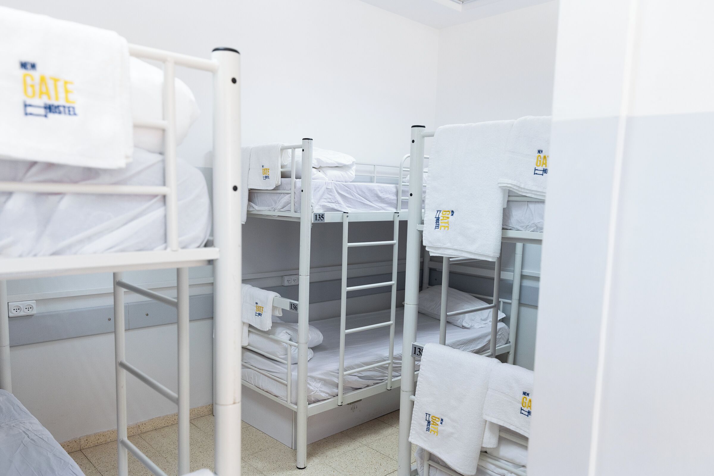 Quarto de dormitório com beliches, cada um com uma toalha branca e uma placa indicando 'New Gate Hostel'.