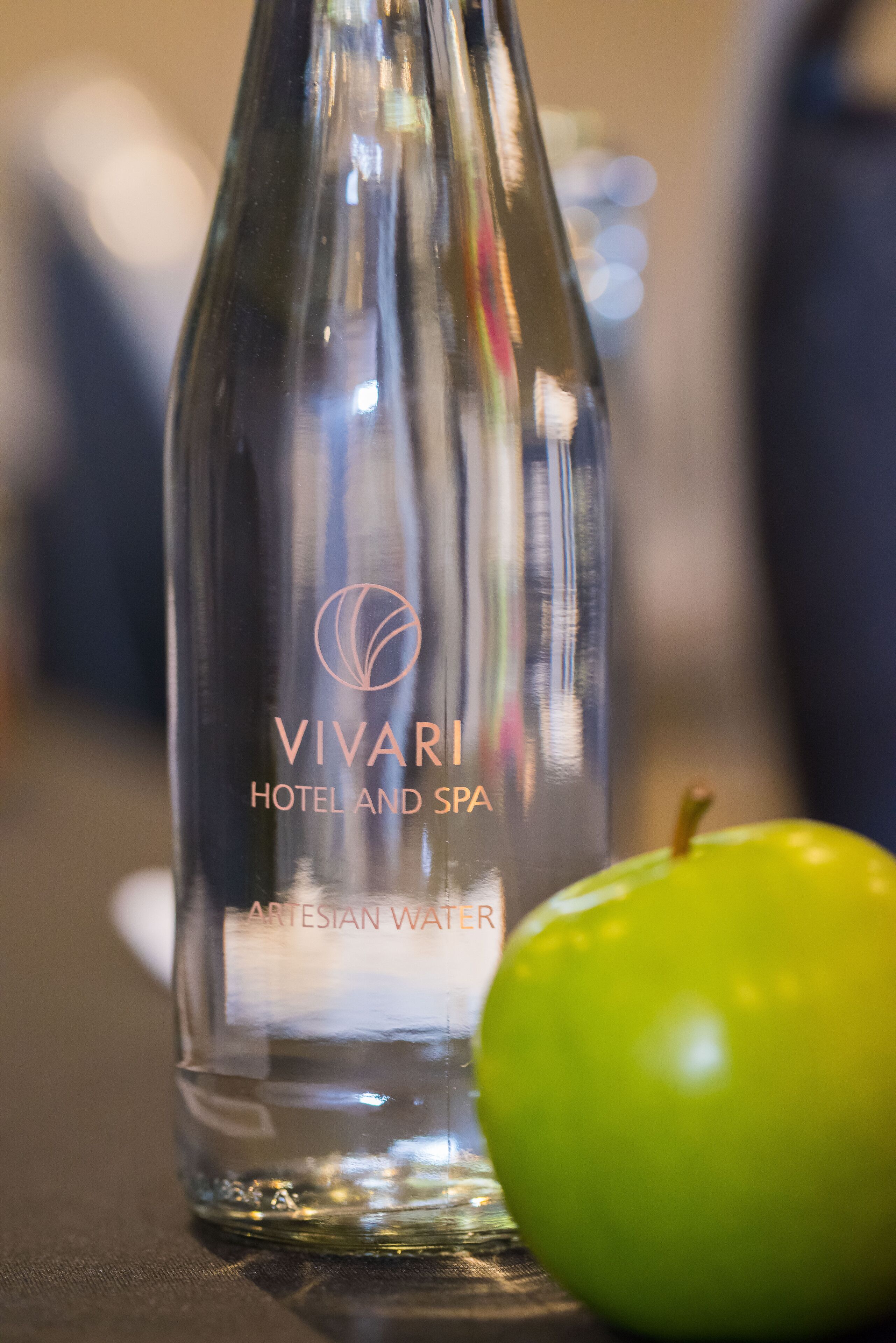 Uma garrafa transparente com o rótulo 'VIVARI HOTEL AND SPA ARTISAN WATER' e uma maçã verde ao lado.