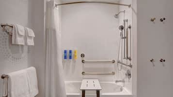 Uma casa de banho moderna com banheira, chuveiro e barras de apoio. Toalhas penduradas num suporte e artigos de higiene pessoal na parede.