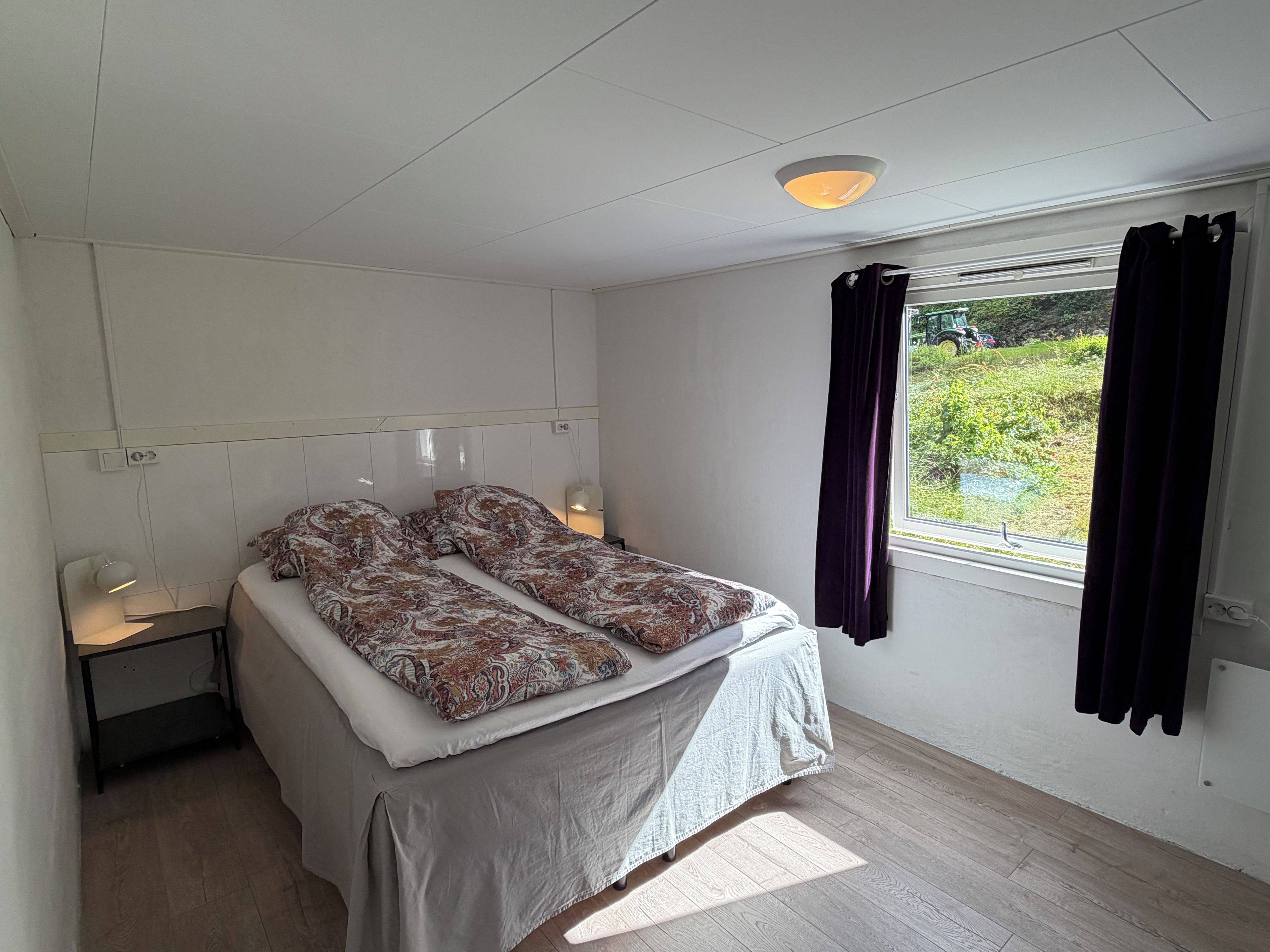 Quarto com cama, abajures, janela com cortinas e vista para o verde lá fora.