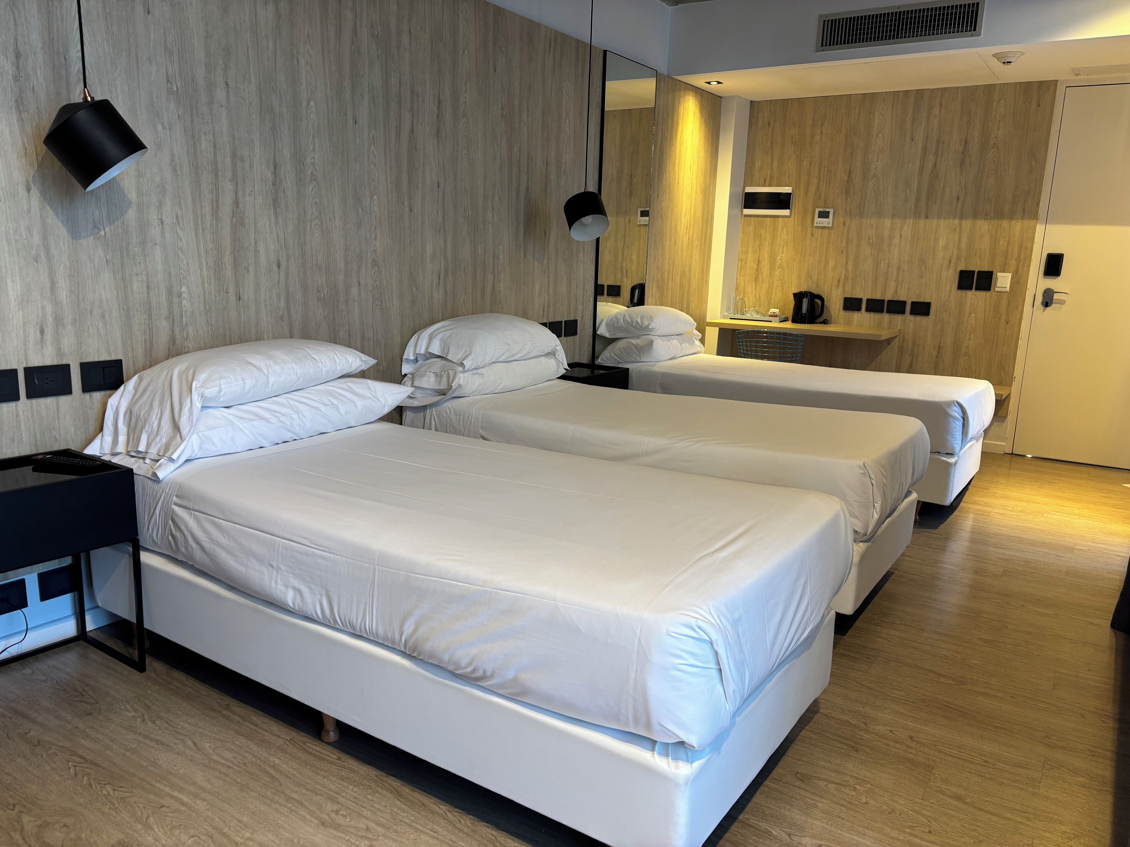 Quarto de hotel com duas camas, uma escrivaninha com luminária, um espelho e parede de madeira.