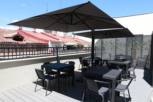 Um pátio externo com mesas e cadeiras, um guarda-sol grande e vista para edifícios com telhados vermelhos.