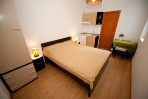 Quarto compacto com cama, mesa de cabeceira com abajur, pequeno espaço de cozinha e porta de madeira.