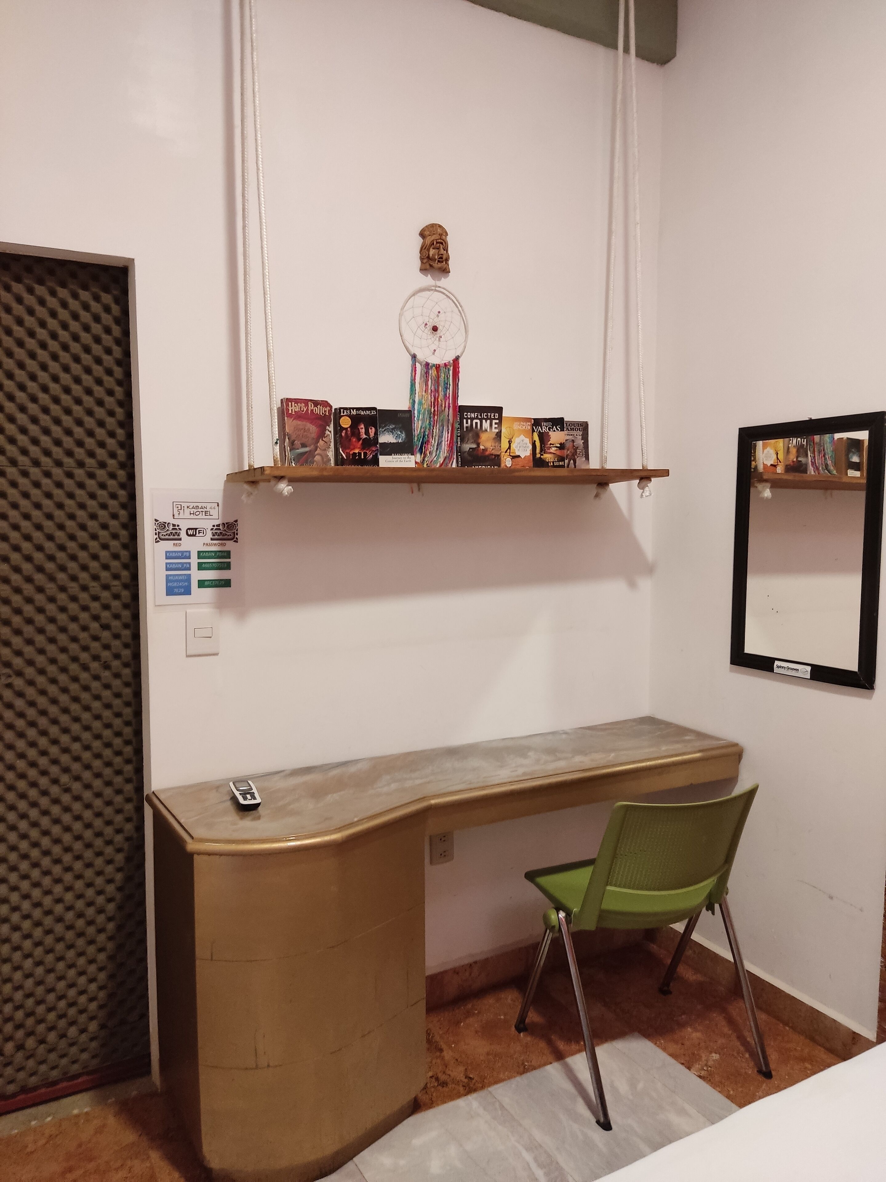 Quarto de hotel com escrivaninha, uma cadeira verde, prateleira com livros, um sonho capturado e uma moldura na parede.