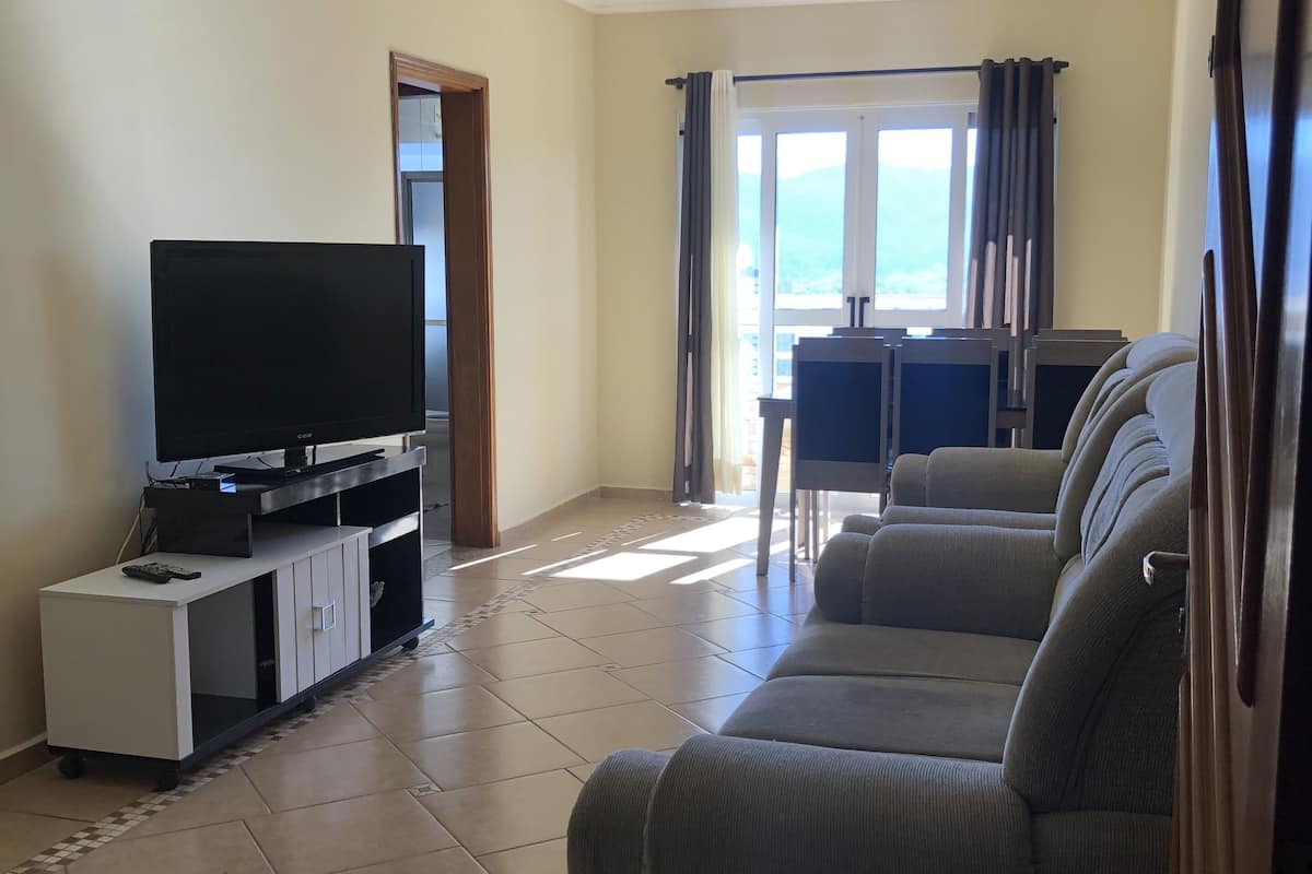 Uma sala de estar com televisão de tela plana em um suporte, um sofá e uma área de jantar com mesa e cadeiras.