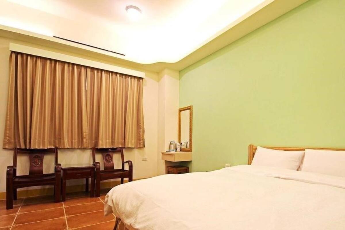 Quarto de hotel com uma cama grande, cabeceira de madeira, mesa de cabeceira, espelho e janela com cortinas.