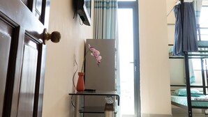 Quarto com porta, ar-condicionado, telefone fixado na parede, planta em vaso, cama e janela com cortinas.