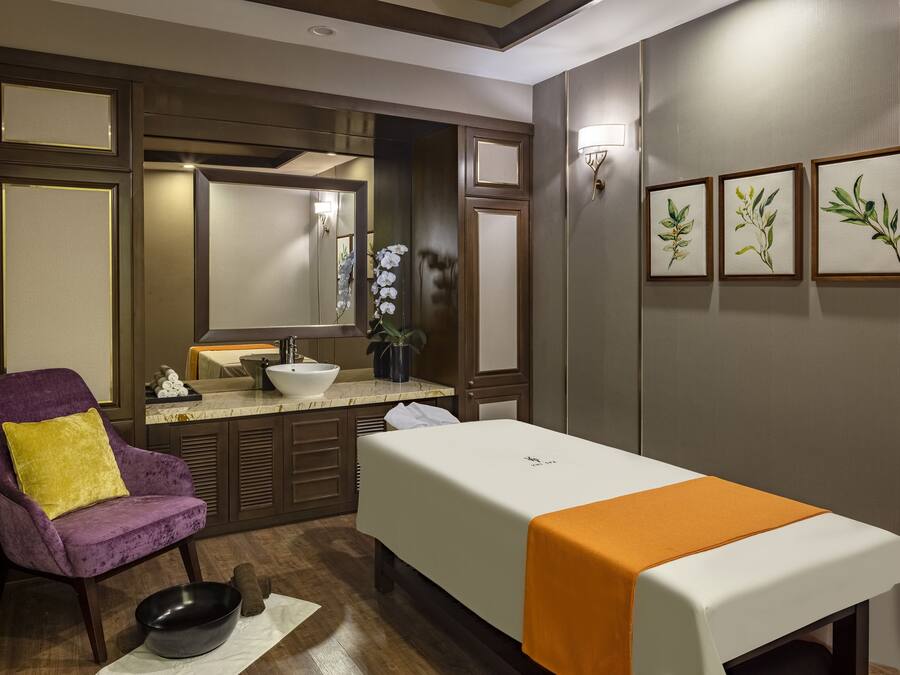 Um quarto com mesa de massagem, uma poltrona roxa, pia e impressões botânicas emolduradas na parede.