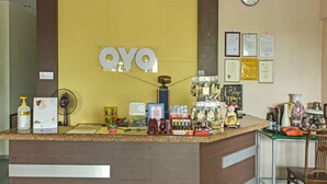 Uma mesa de recepção com uma placa indicando "OYO" e diversos objetos, como um ventilador, artigos de higiene pessoal e uma pequena estátua.