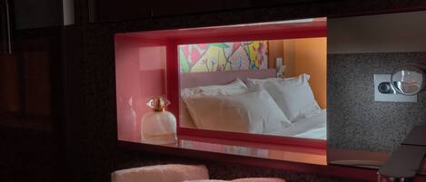 Quarto de hotel com cama, espelho, mesinha e um objeto decorativo na parede.