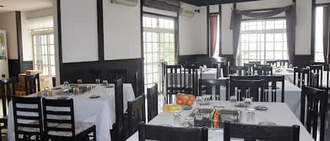 Uma sala de jantar com várias mesas e cadeiras, grandes janelas com cortinas e toalhas de mesa brancas.