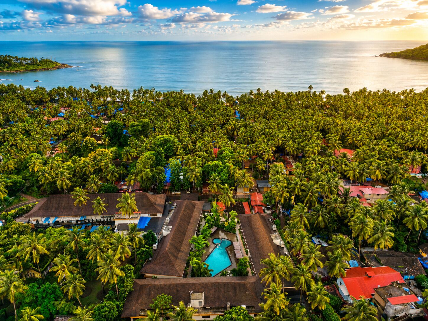 Vista aérea de um resort com piscina, cercado por palmeiras e edifícios com telhados vermelhos.