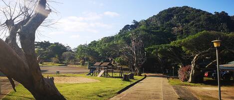 Um parque com um parquinho, uma trilha pavimentada e uma colina ao fundo.