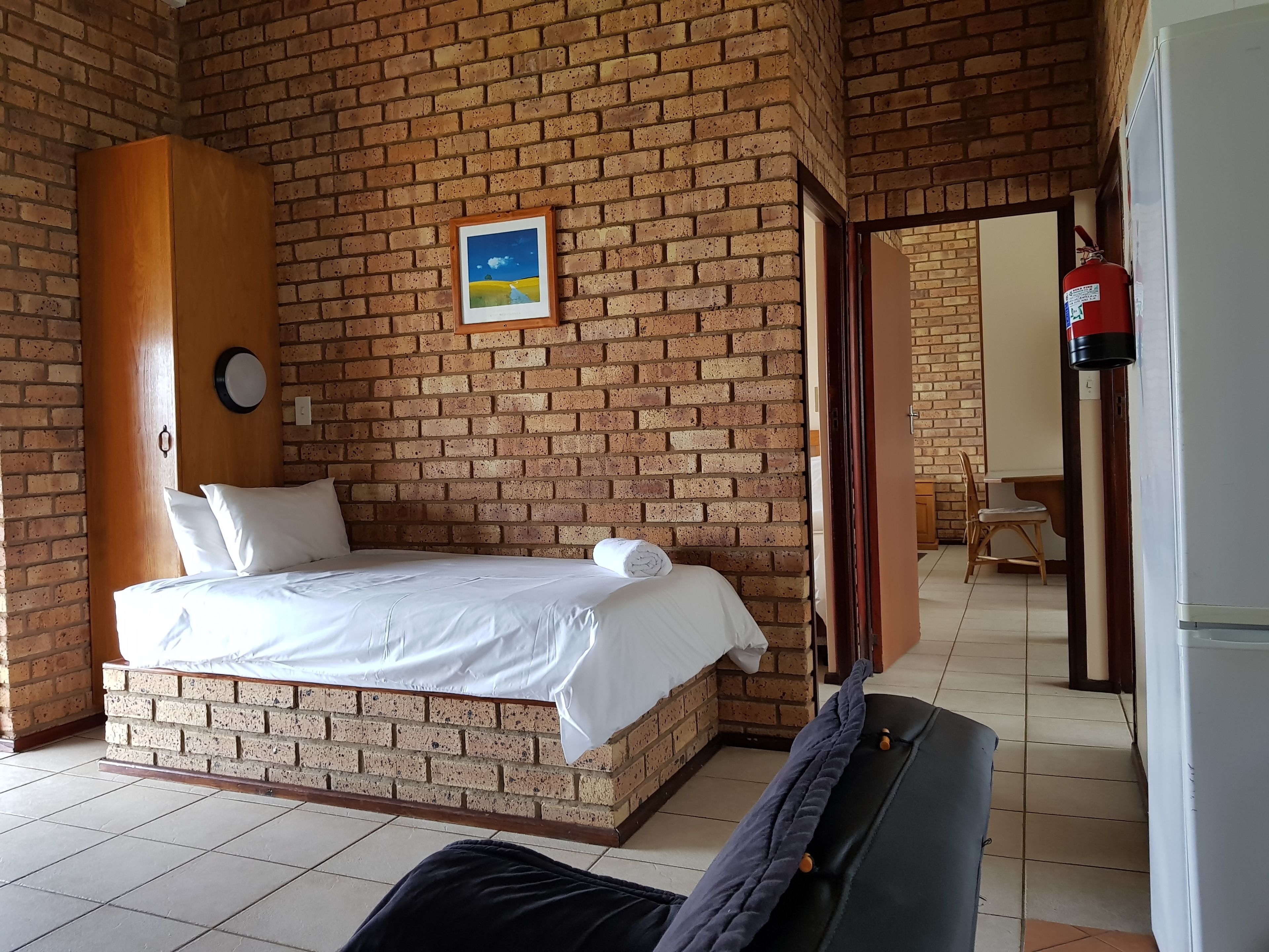 Quarto com parede de tijolos, cama com roupa de cama branca, espelho redondo e quadro na parede.