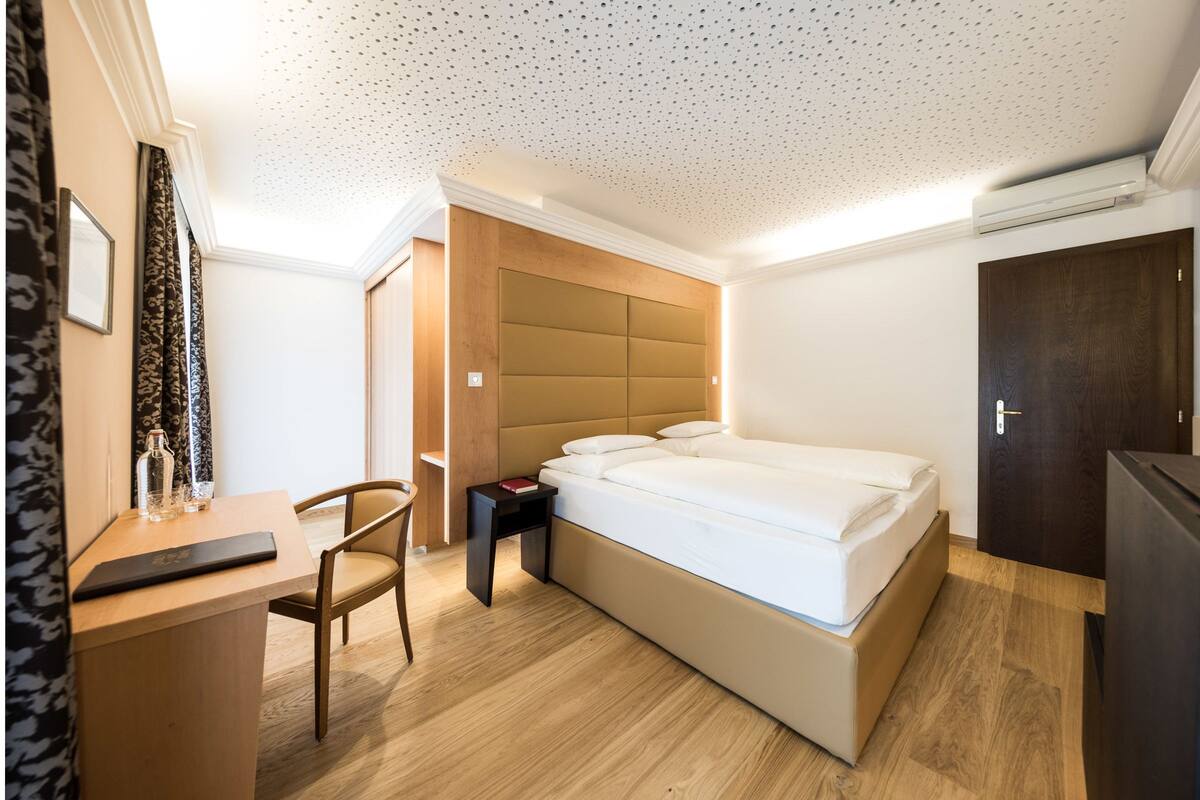 Quarto de hotel moderno com uma cama grande, uma escrivaninha com cadeira, uma mesinha com uma garrafa e copos, e um guarda-roupa.