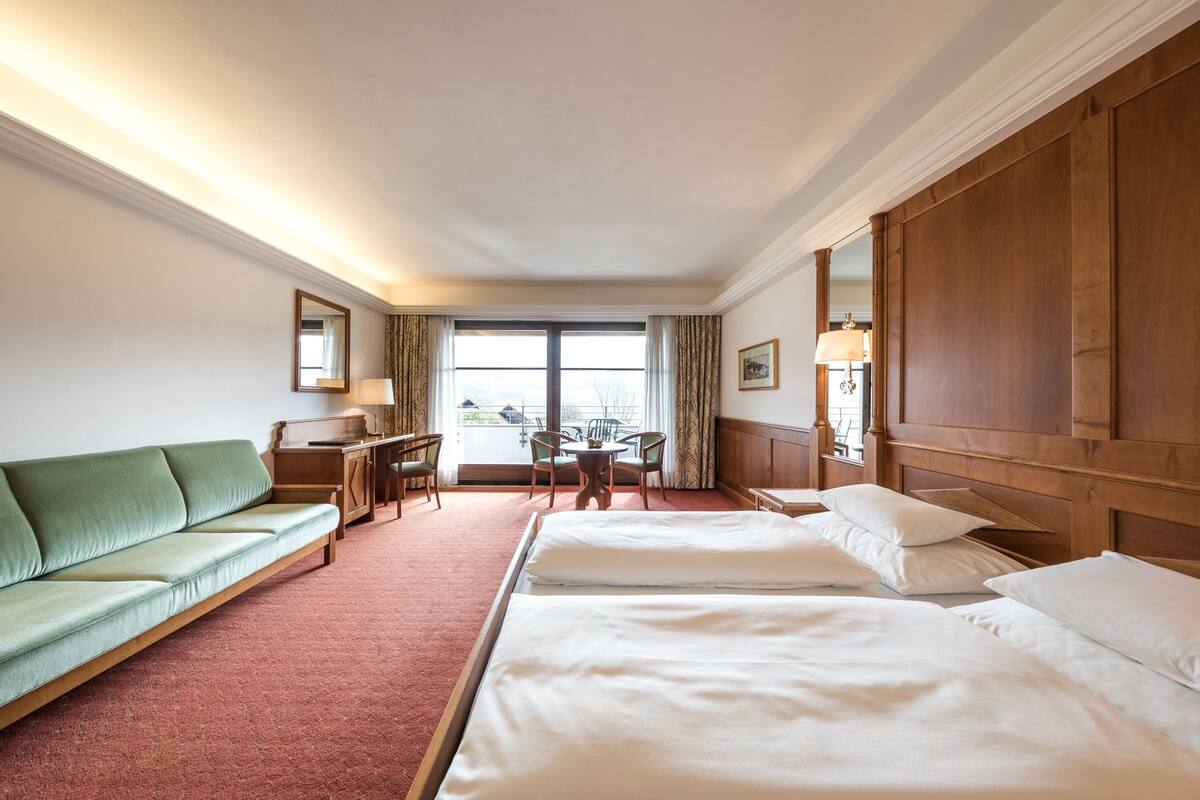 Quarto de hotel com um sofá verde, uma escrivaninha com cadeiras, duas camas e uma janela grande com cortinas.