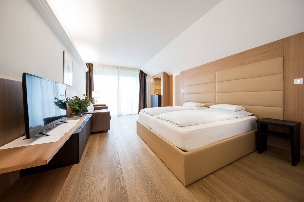 Quarto de hotel moderno com uma cama grande, uma escrivaninha com televisão de tela plana, um pequeno espaço para sentar e uma janela ampla com cortinas.