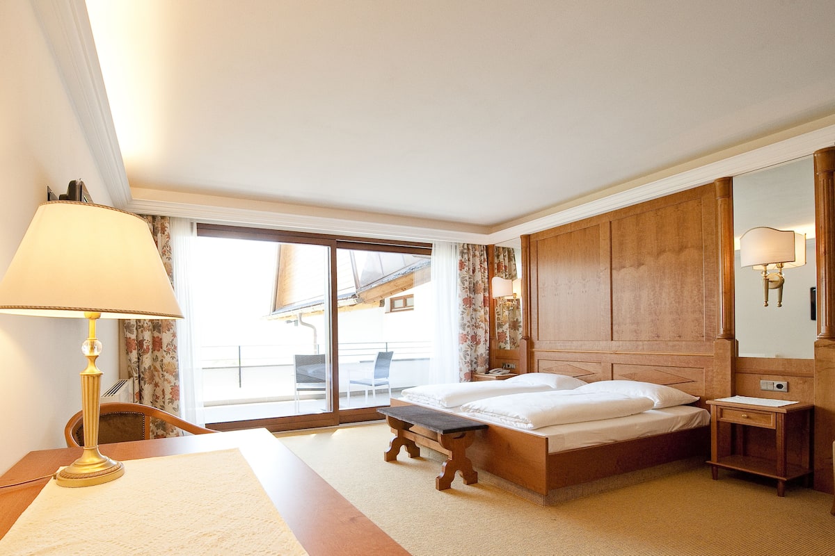 Quarto de hotel com cama, mesa de cabeceira, luminária e uma grande janela com vista para o exterior.