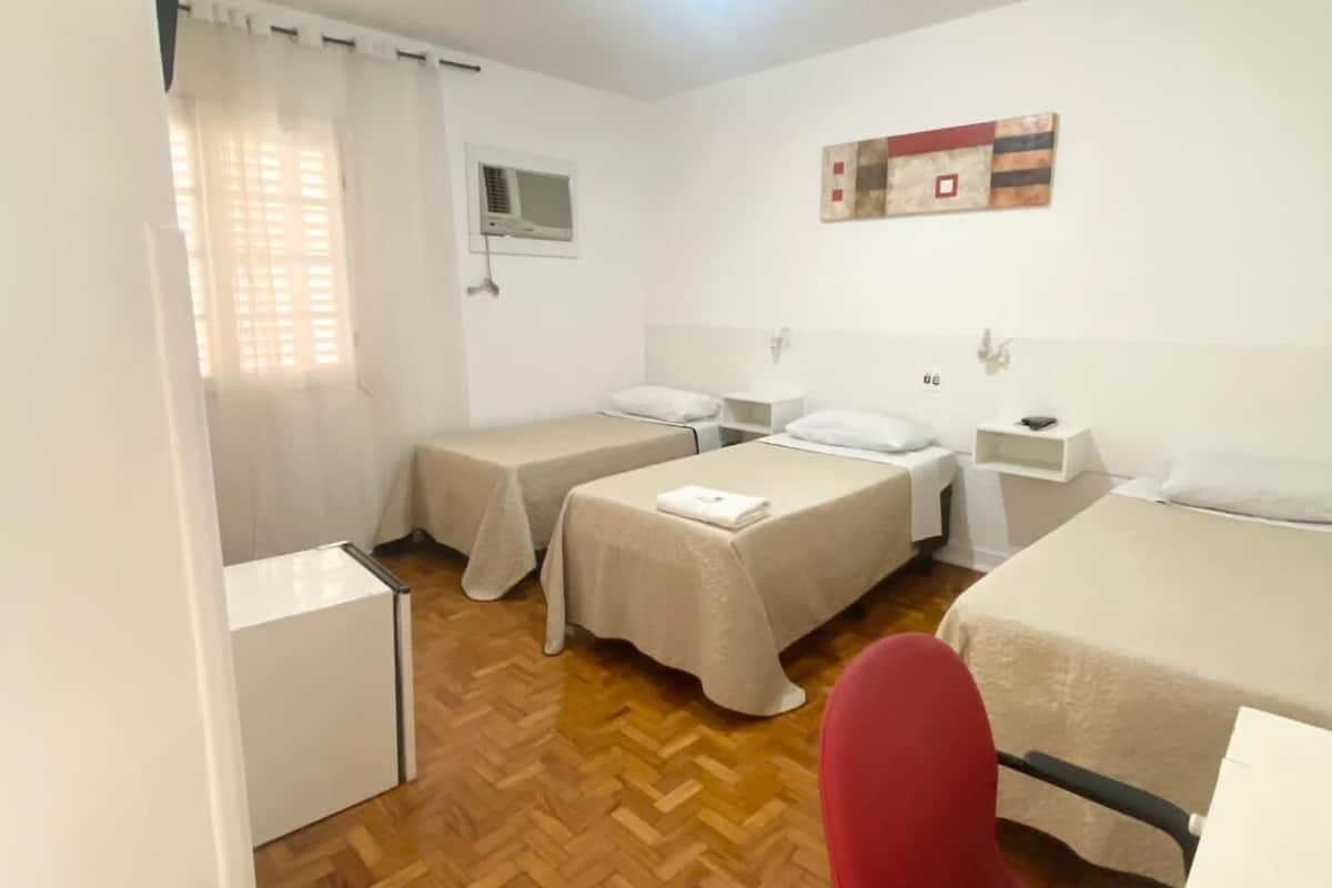 Quarto de hotel com duas camas de solteiro, uma escrivaninha pequena, uma cadeira e uma janela com cortinas.
