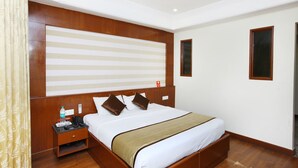 Quarto de hotel com cabeceira de madeira, uma janela grande com persianas, mesa de cabeceira com telefone e garrafa, e cama com roupa de cama branca e almofadas castanhas.