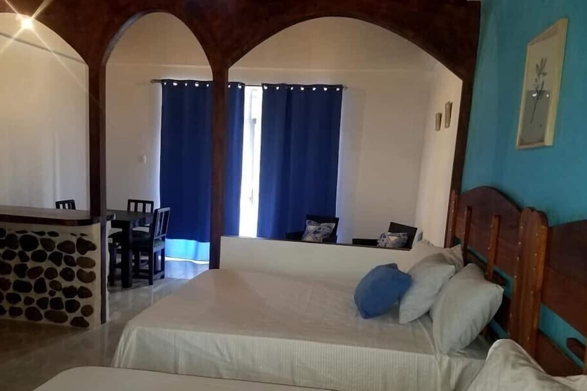 Quarto com cama, uma pequena mesa e uma cadeira. Há uma janela com cortinas e um relógio na parede.