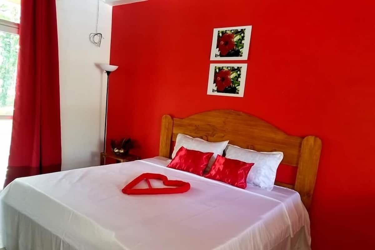 Cama com roupa de cama branca e almofadas vermelhas, cabeceira de madeira e duas imagens de flores vermelhas na parede.