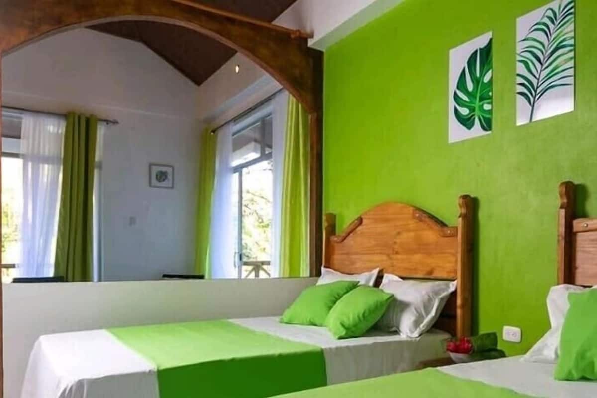 Quarto com duas camas, roupa de cama verde, cabeceiras de madeira e arte na parede com padrão de folhas.