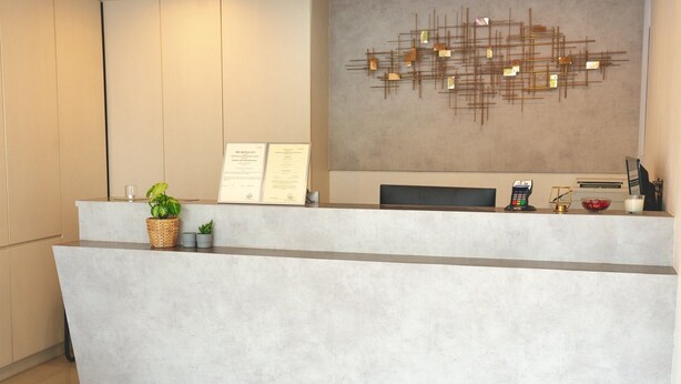 Balcão de recepção de hotel com design moderno, uma obra de arte abstrata fixada na parede, um certificado emoldurado, uma planta, uma pequena planta em vaso, um terminal de pagamento e uma vela.