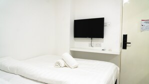 Quarto de hotel com uma cama, uma TV de tela plana na parede, uma porta com fechadura e uma pequena estante com alguns objetos.