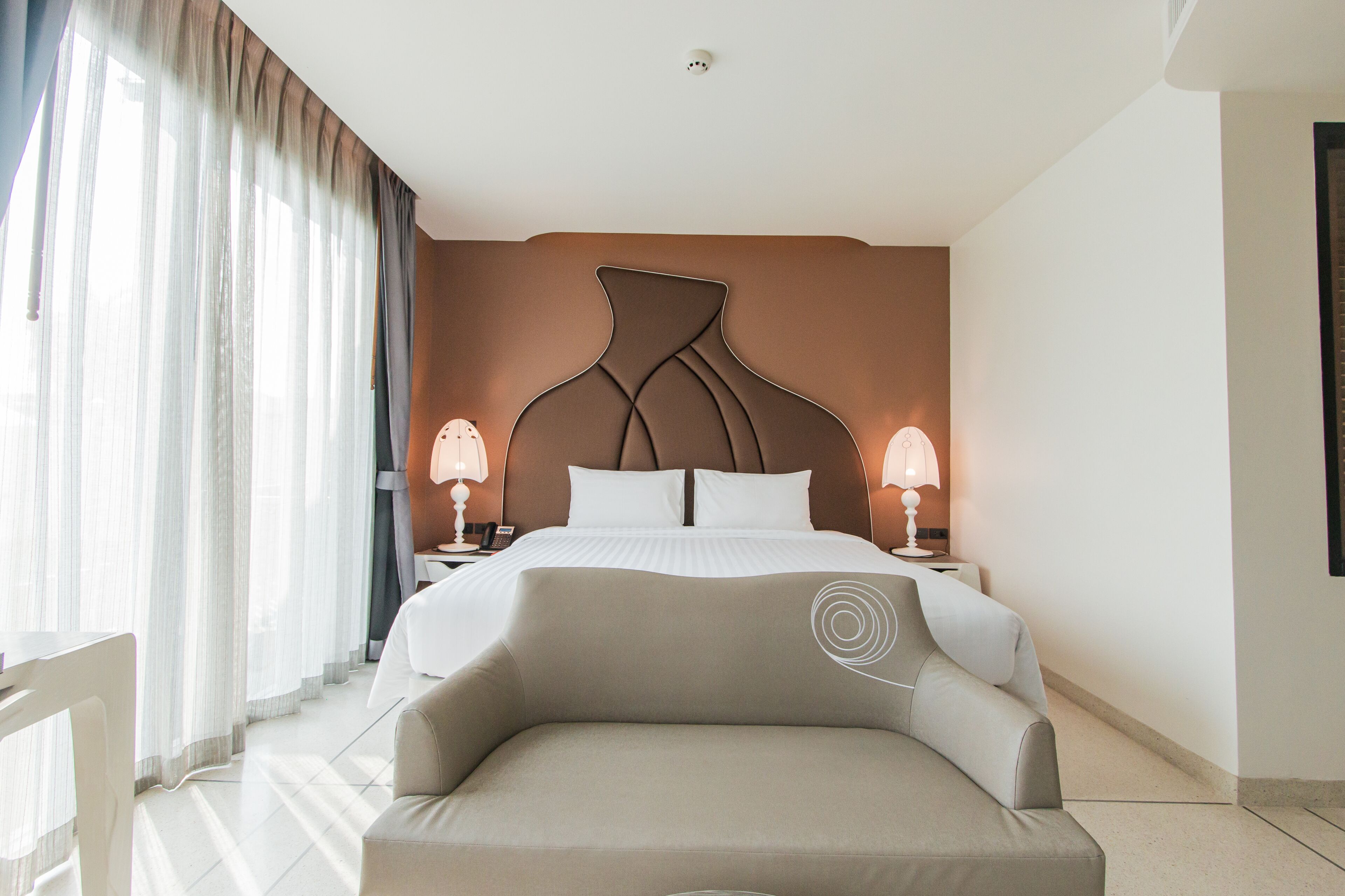 Quarto de hotel moderno com uma cama grande, duas luminárias laterais, um sofá e cortinas translúcidas.