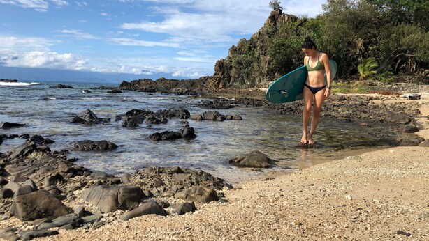 Uma praia rochosa com águas cristalinas, uma pessoa segurando uma prancha de surf e um cenário montanhoso ao fundo.