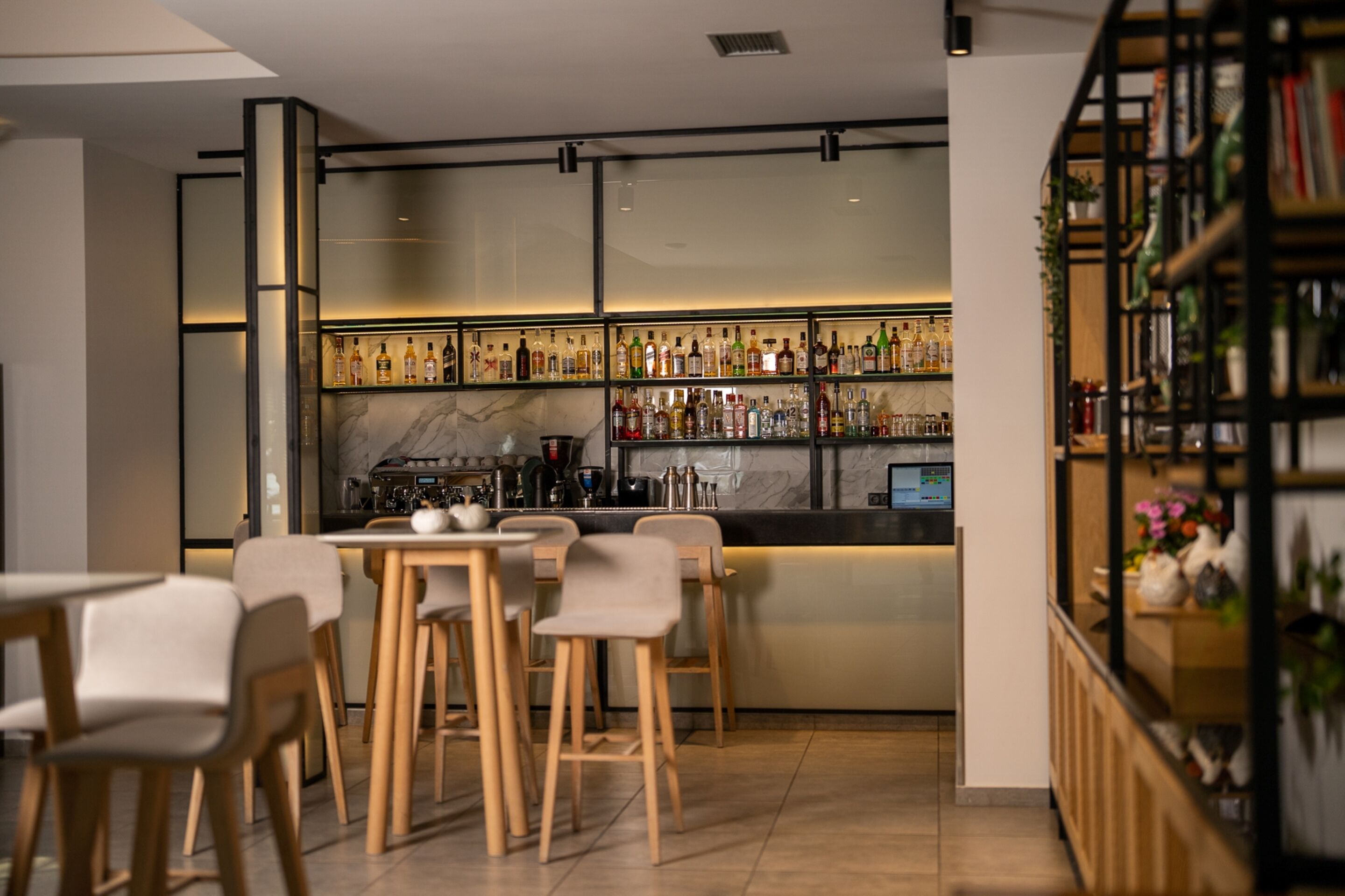 Uma área de bar moderna com bancada de mármore, várias prateleiras com garrafas de bebidas e uma máquina de café. Cadeiras de madeira e uma pequena mesa estão em primeiro plano.