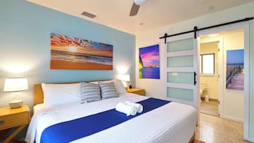 Quarto de hotel com uma cama, duas mesas de cabeceira, um ventilador de teto e vista para a praia pela porta.