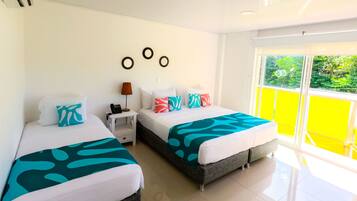 Quarto de hotel com duas camas, uma pequena mesa de cabeceira com abajur e uma janela grande com vista para o verde.