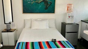 Quarto de hotel com cama, manta listrada colorida, pintura de um mergulhador, bebedouro e escrivaninha com luminária.
