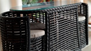 Mesa de vime preta com assentos acolchoados, posicionada sobre uma mesa de vidro em um quarto com vista para o verde através de grandes janelas.