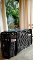 Mesa de vime preta com assentos acolchoados, posicionada sobre uma mesa de vidro em um quarto com vista para o verde através de grandes janelas.