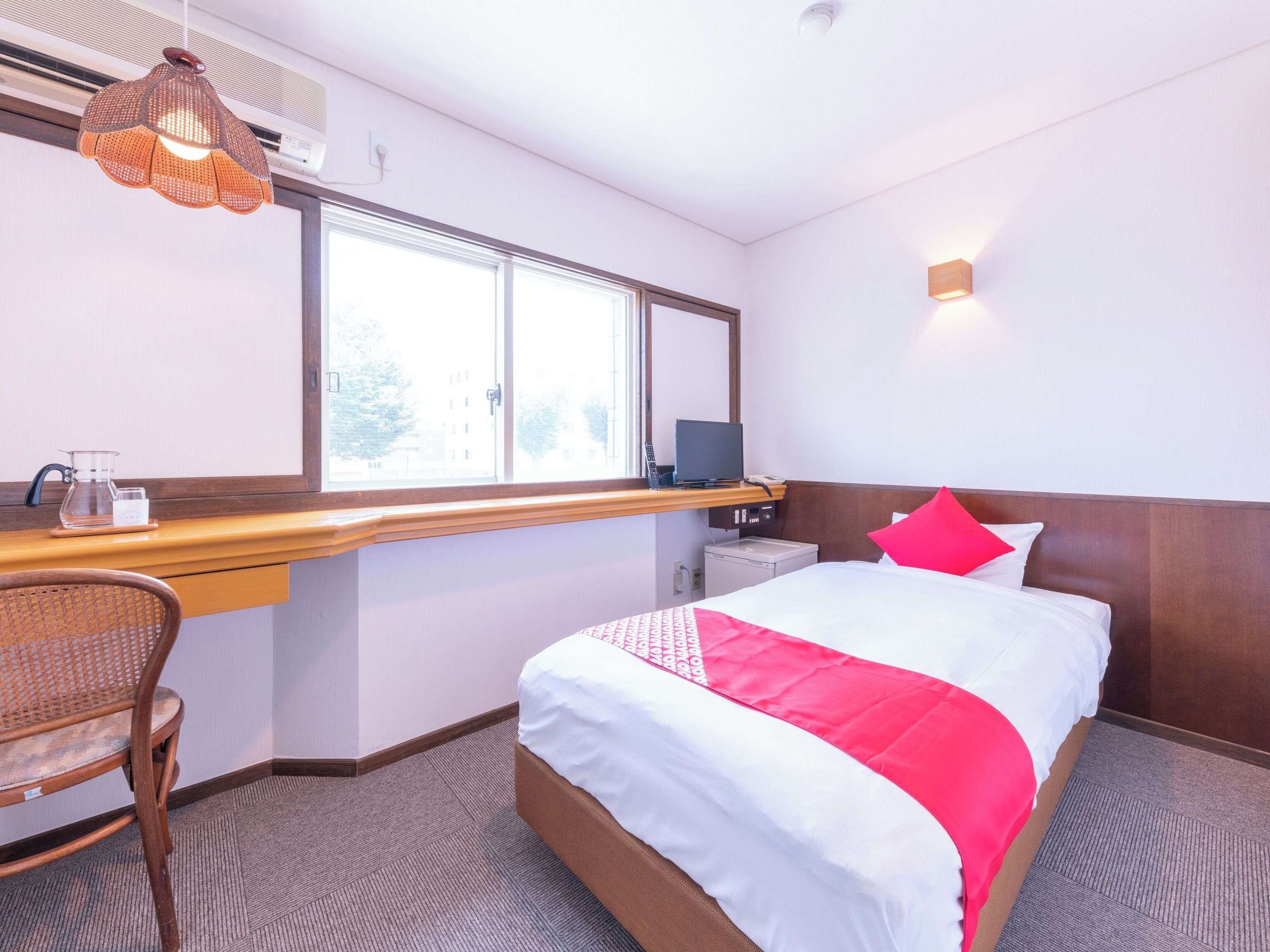 Quarto de hotel com cama, escrivaninha, cadeira, janela e luminária fixada na parede.