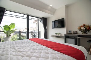 Quarto de hotel com uma cama grande, televisão presa na parede, escrivaninha com um computador e varanda com uma planta.