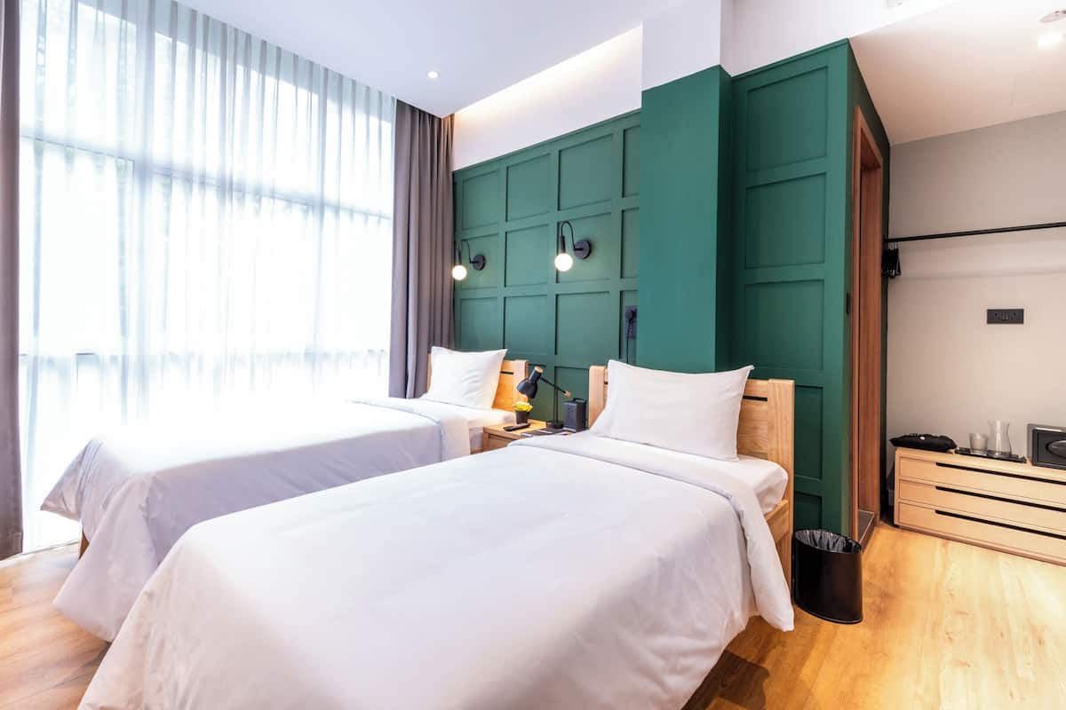 Quarto de hotel com duas camas, parede de destaque verde, chão de madeira e janela com cortinas.