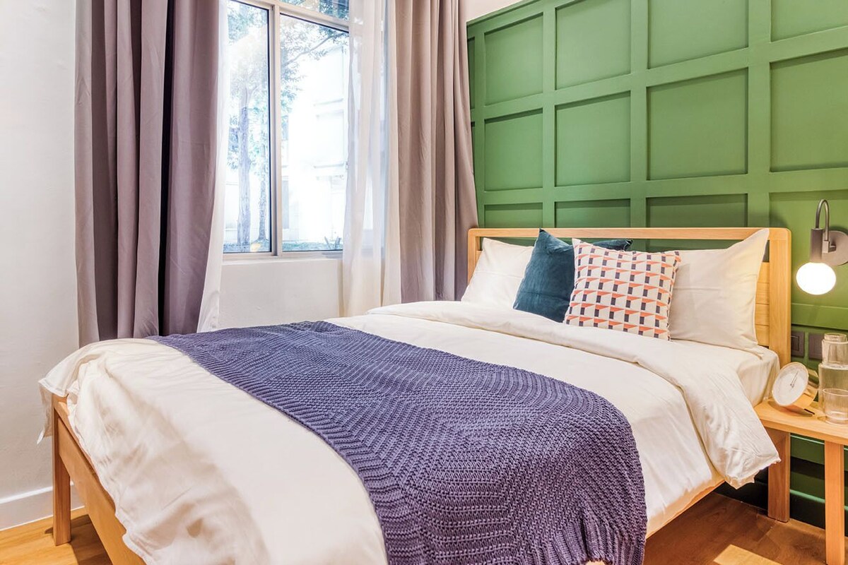 Uma cama bem arrumada com um edredom branco e azul, cabeceira verde e mesa de cabeceira com abajur e relógio.