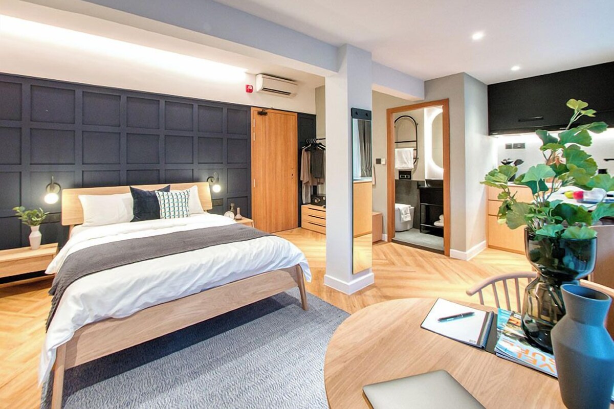 Quarto de hotel moderno com uma cama grande, uma mesa de cabeceira, uma escrivaninha com cadeira, um espelho, um guarda-roupa e um banheiro visível através de uma porta aberta.