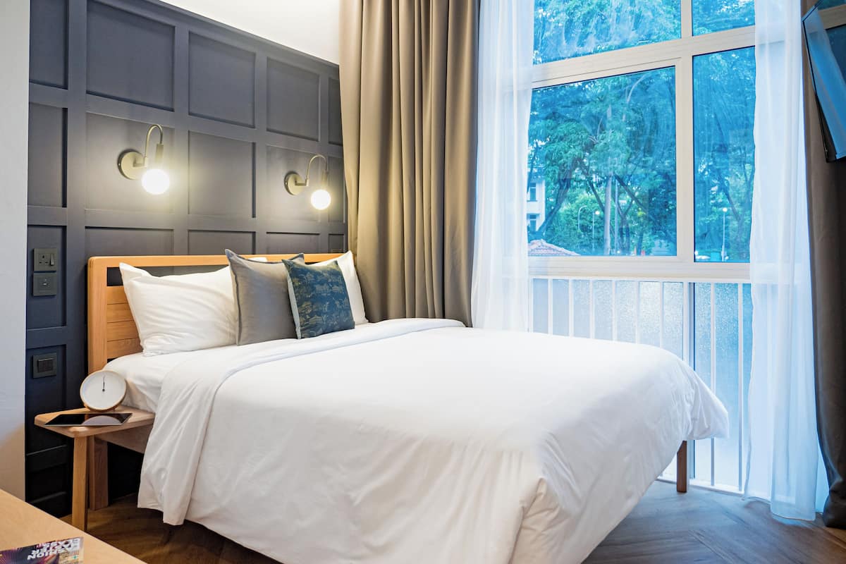 Cama bem arrumada com roupa de cama branca e duas almofadas, mesa de cabeceira com relógio e tablet, e janela com cortinas.