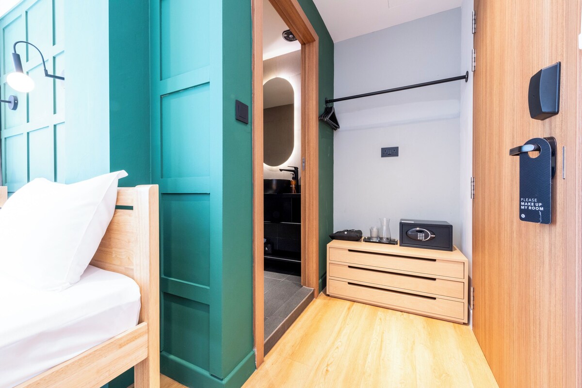 Quarto de hotel pequeno com uma cama, cômoda de madeira, espelho e porta com uma placa.